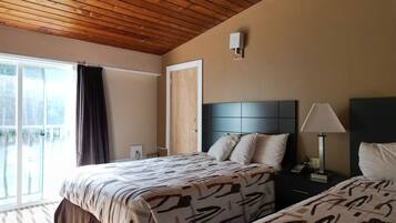 Quarto de hotel com teto de madeira, uma janela grande com cortinas, duas camas com roupa de cama estampada, uma mesa de cabeceira com abajur e um candeeiro fixado na parede.