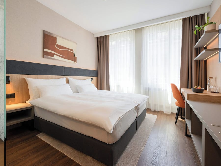 Quarto de hotel com cama, escrivaninha, cadeira e televisão.