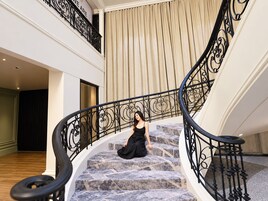 Mulher sentada em escadaria curva com corrimão preto, que leva a um andar superior.