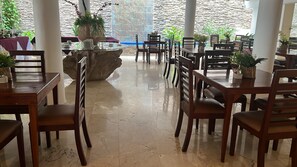 Uma ampla área de refeições interna com piso de mármore, mesas e cadeiras de madeira e plantas em vasos.