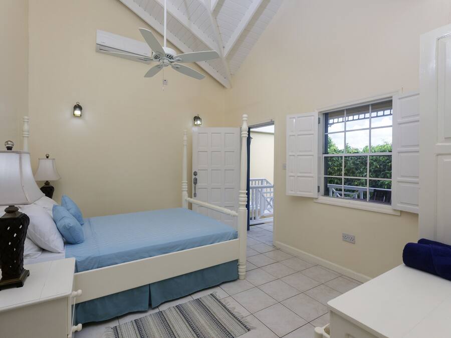 Quarto com cama, mesa de cabeceira, abajur, ventoinha no teto e janela com persianas.