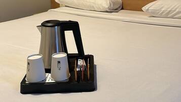 Quarto de hotel com cabeceira de madeira, cama com lençóis brancos, duas almofadas, abajur e bandeja com cafeteira, duas chávenas e um termo.