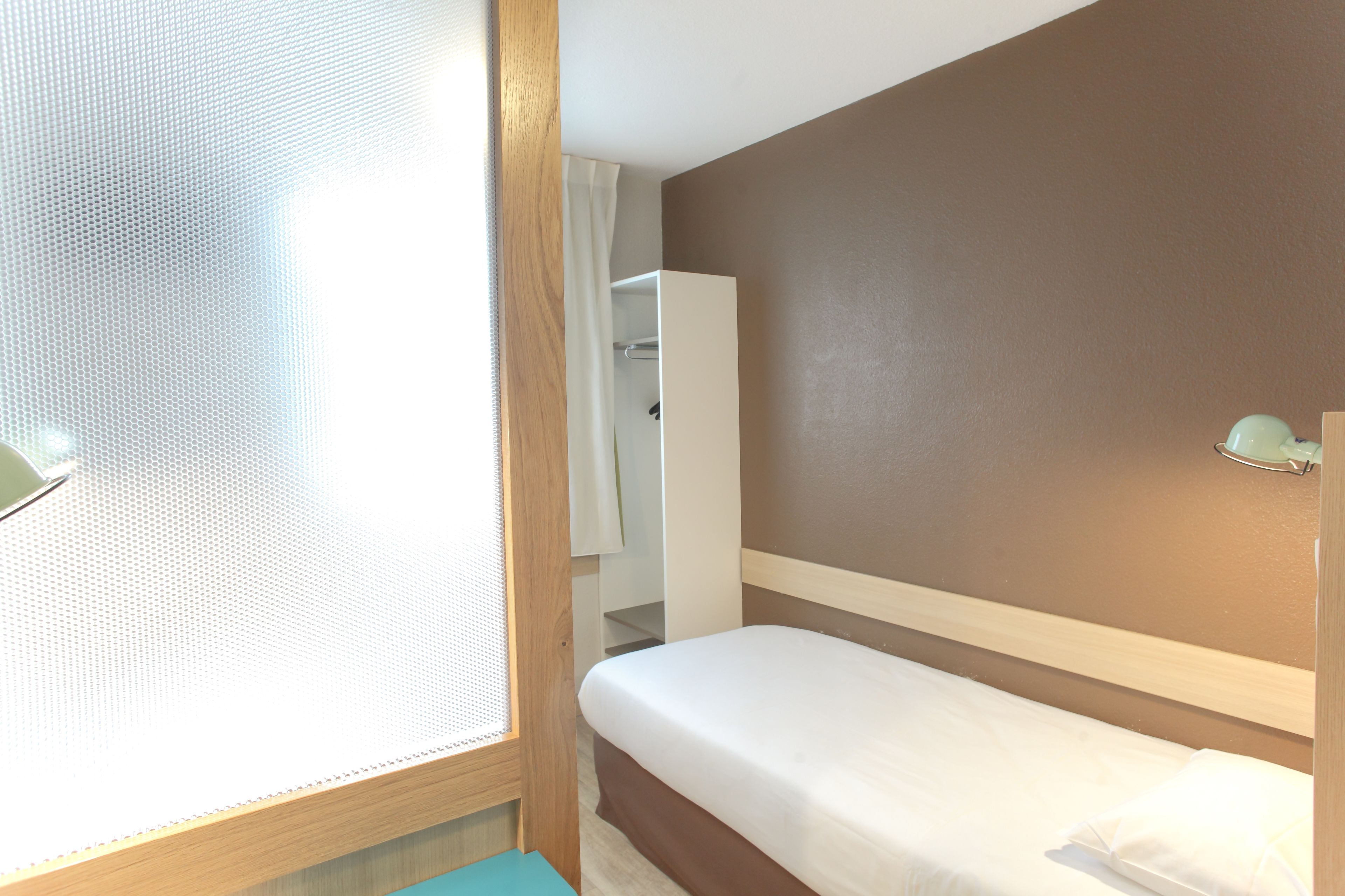 Quarto pequeno com cama de solteiro, cabeceira de madeira, janela com cortina branca e guarda-roupa branco ao fundo.