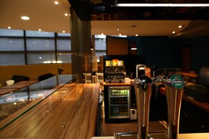 Uma área de bar com balcão de madeira, divisória de vidro e geladeira abastecida com bebidas.