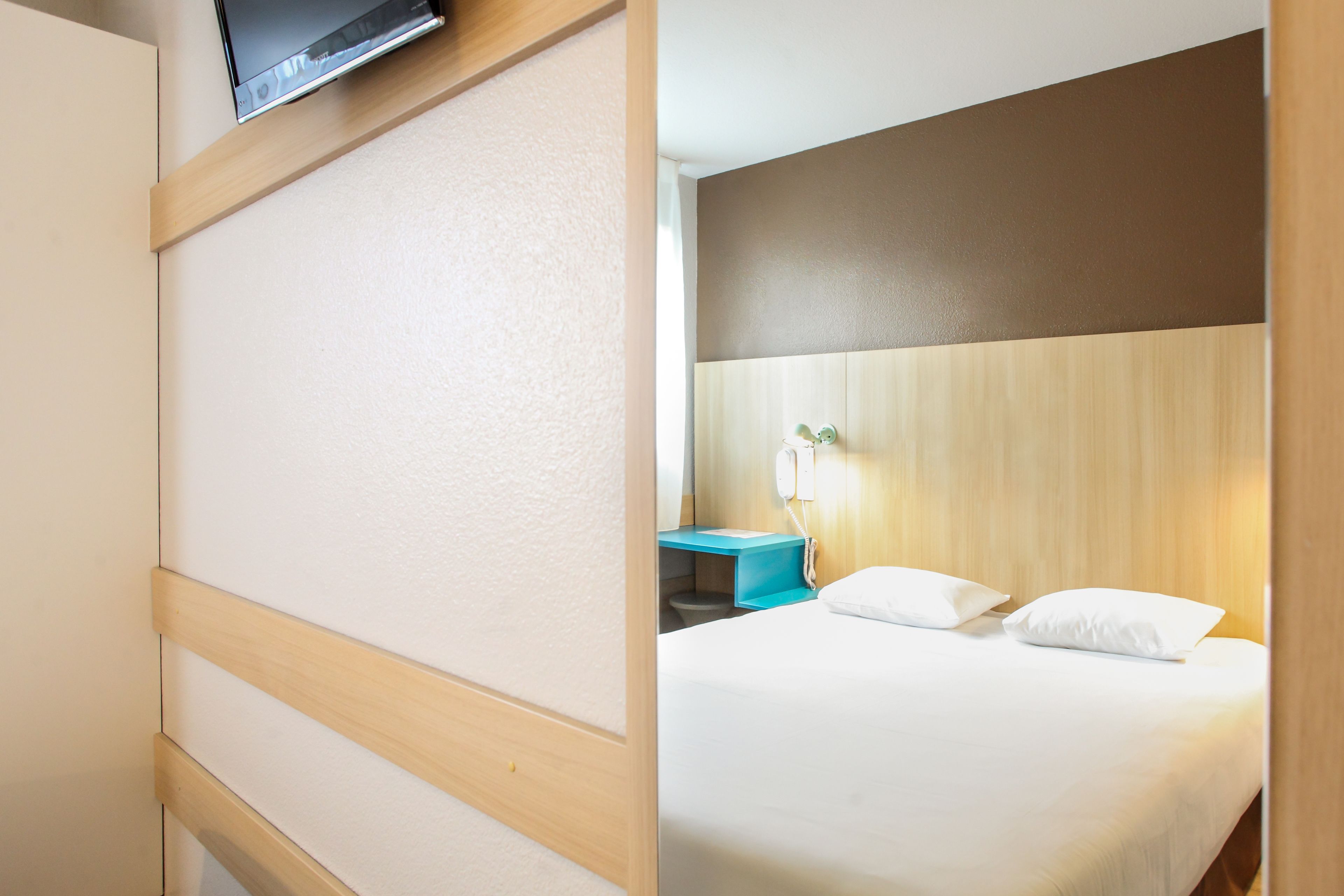 Quarto de hotel com uma cama, mesa de cabeceira, telefone e televisão fixada na parede.
