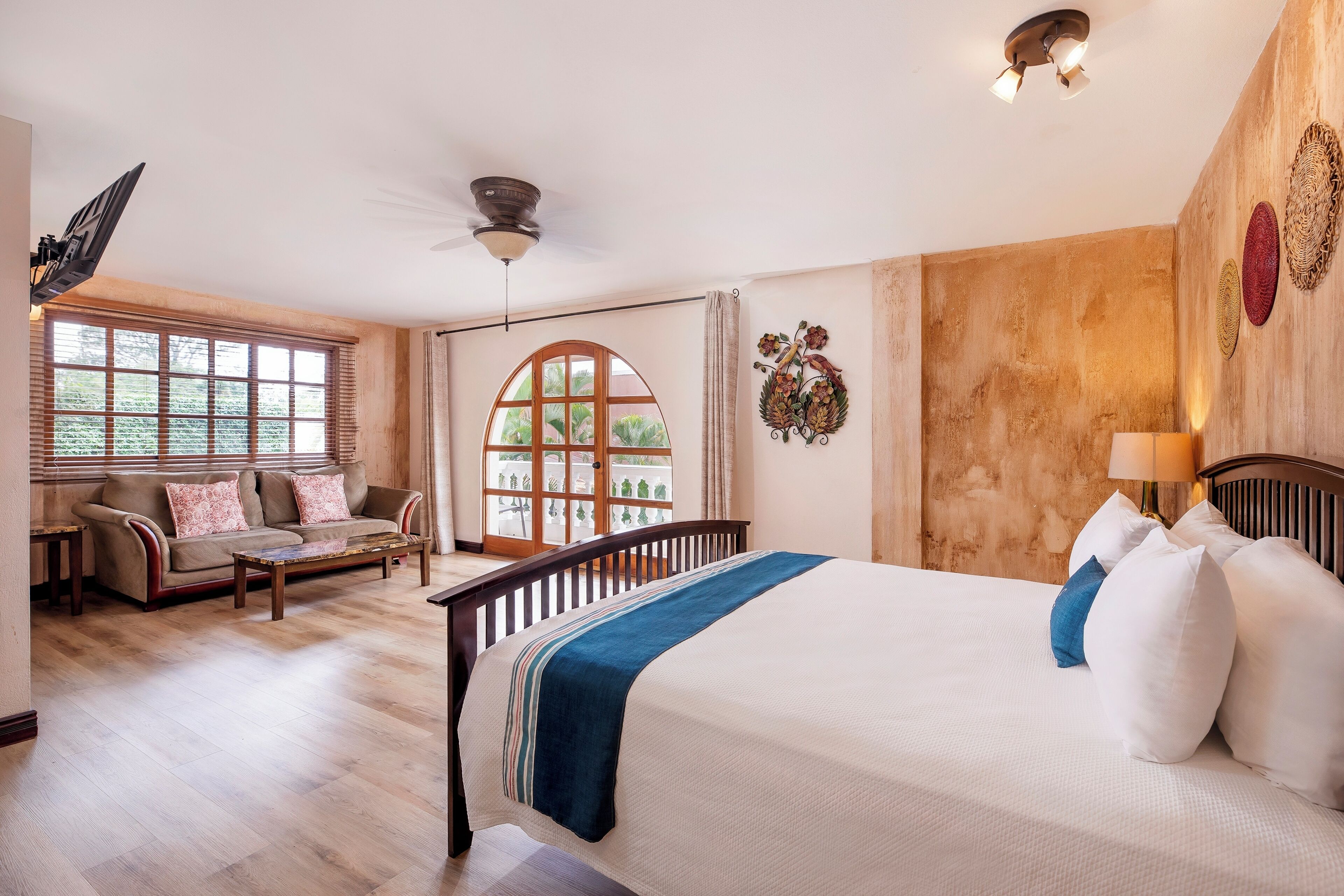 Quarto de hotel com cama, sofá, mesa de centro, ventoinha no teto e janela com cortinas.