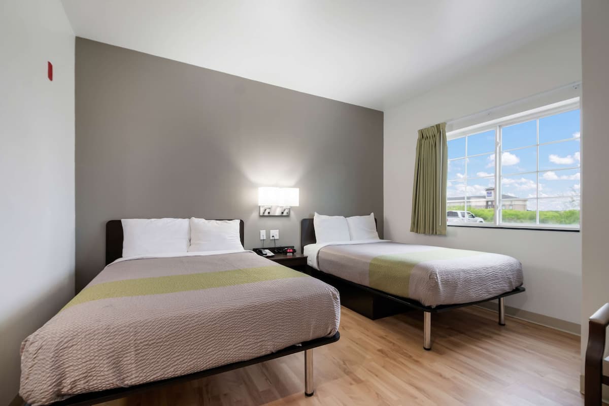 Quarto de hotel com duas camas, uma mesa de cabeceira, uma luminária e uma janela com vista para o céu e prédios.