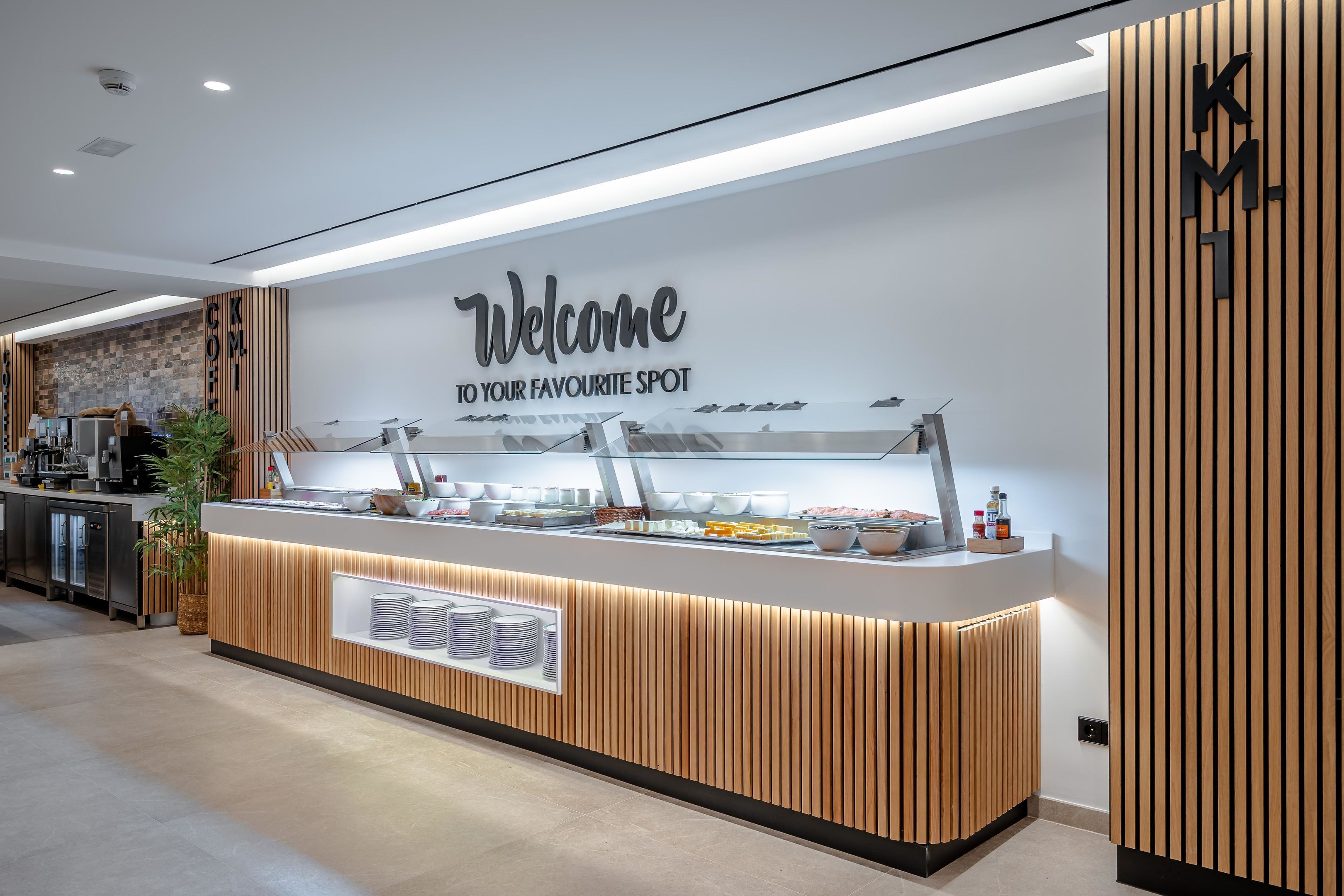 Uma área de recepção moderna com um aviso 'Bem-vindo ao seu lugar preferido', um balcão de buffet com diversas opções de comida e painéis de madeira nas paredes.