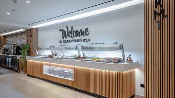 Uma área de recepção moderna com um aviso 'Bem-vindo ao seu lugar preferido', um balcão de buffet com diversas opções de comida e painéis de madeira nas paredes.
