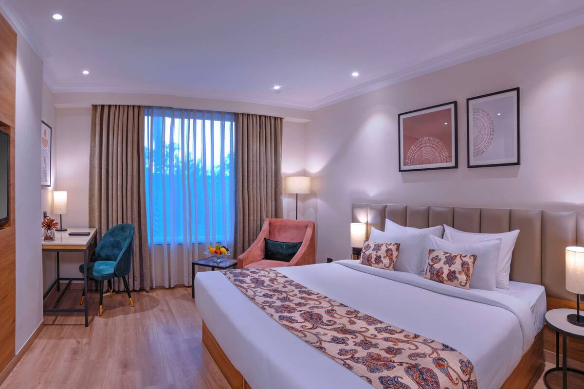 Quarto de hotel com cama, escrivaninha, cadeira, televisão e janela com cortinas.