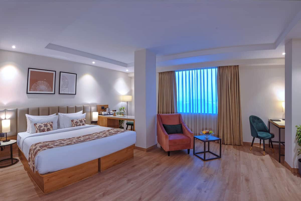 Quarto de hotel com uma cama grande, uma escrivaninha, duas cadeiras, uma mesa pequena e uma planta em vaso.