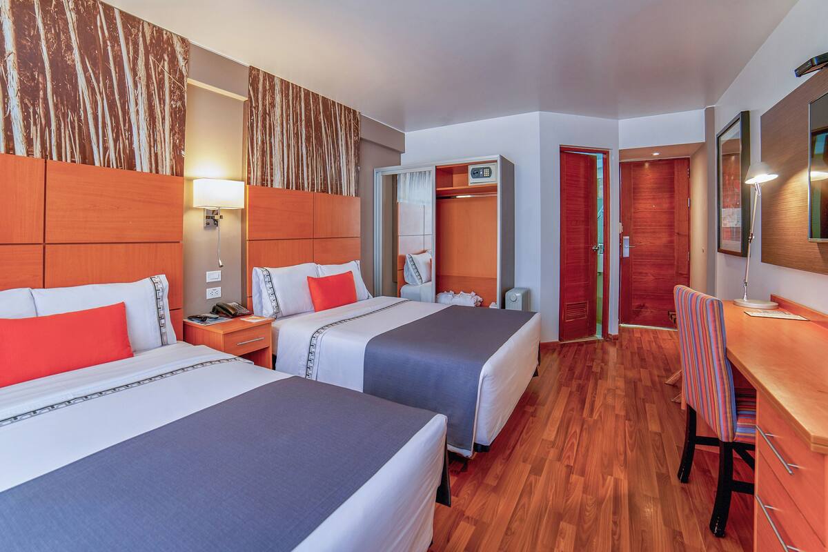 Quarto de hotel com duas camas, uma escrivaninha, uma cadeira, um guarda-roupa e um espelho.