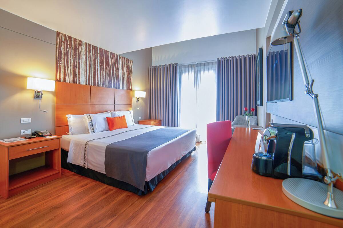 Quarto de hotel com cama, mesa de cabeceira, luminária, escrivaninha, cadeira e máquina de café.