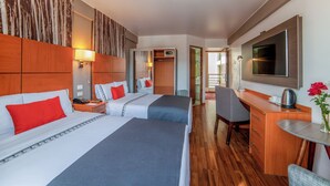 Quarto de hotel com duas camas, uma escrivaninha, uma cadeira, uma televisão e um banheiro visível por uma porta aberta.