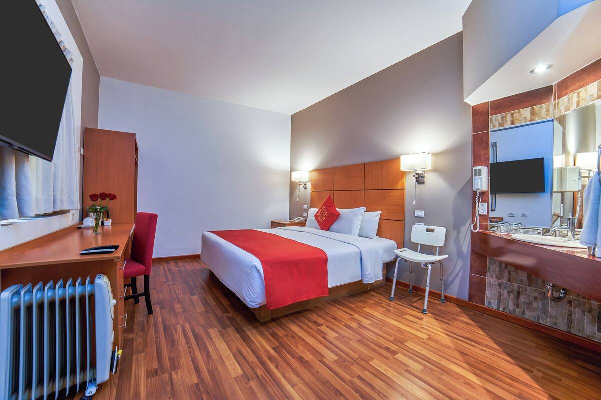 Quarto de hotel com cama, escrivaninha, cadeira, radiador, televisão e banheiro com espelho e pia.