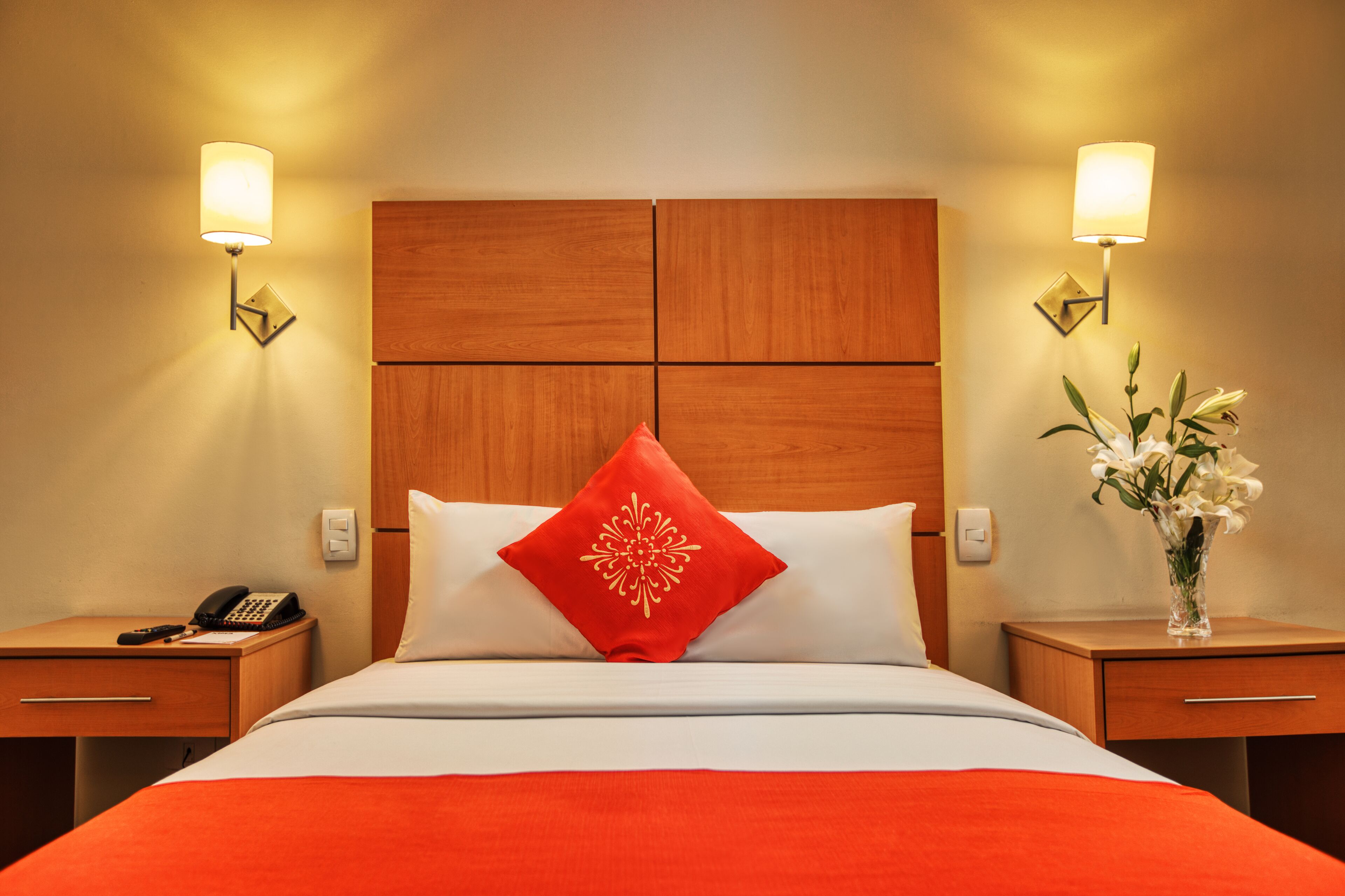 Quarto de hotel com cabeceira de madeira, travesseiro decorativo vermelho, telefone ao lado da cama e um vaso com flores na mesinha de cabeceira.