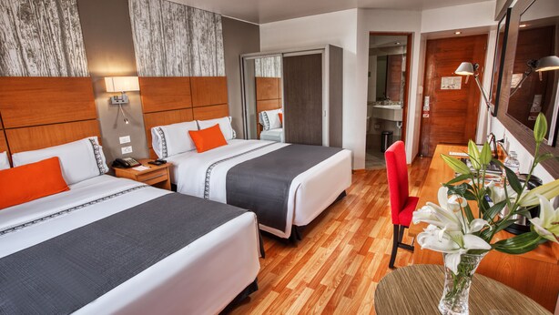 Quarto de hotel com duas camas, uma escrivaninha, uma cadeira, um espelho e um vaso com flores.