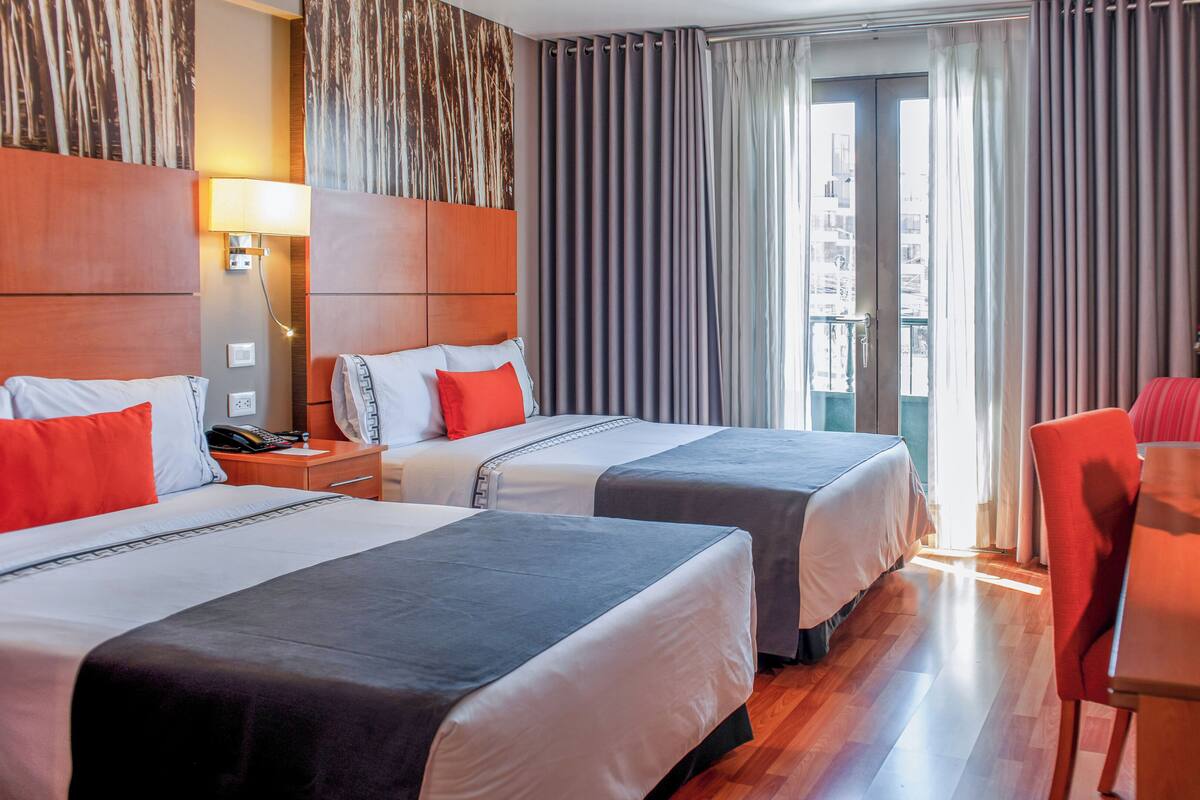 Quarto de hotel com duas camas, uma escrivaninha, uma cadeira, um abajur e uma janela com cortinas.