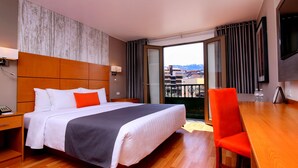 Quarto de hotel com cama, escrivaninha, cadeira, televisão e varanda com vista para edifícios.