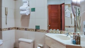 Casa de banho com sanita, lavatório e espelho. Toalhas penduradas na parede e um secador de cabelo no balcão.