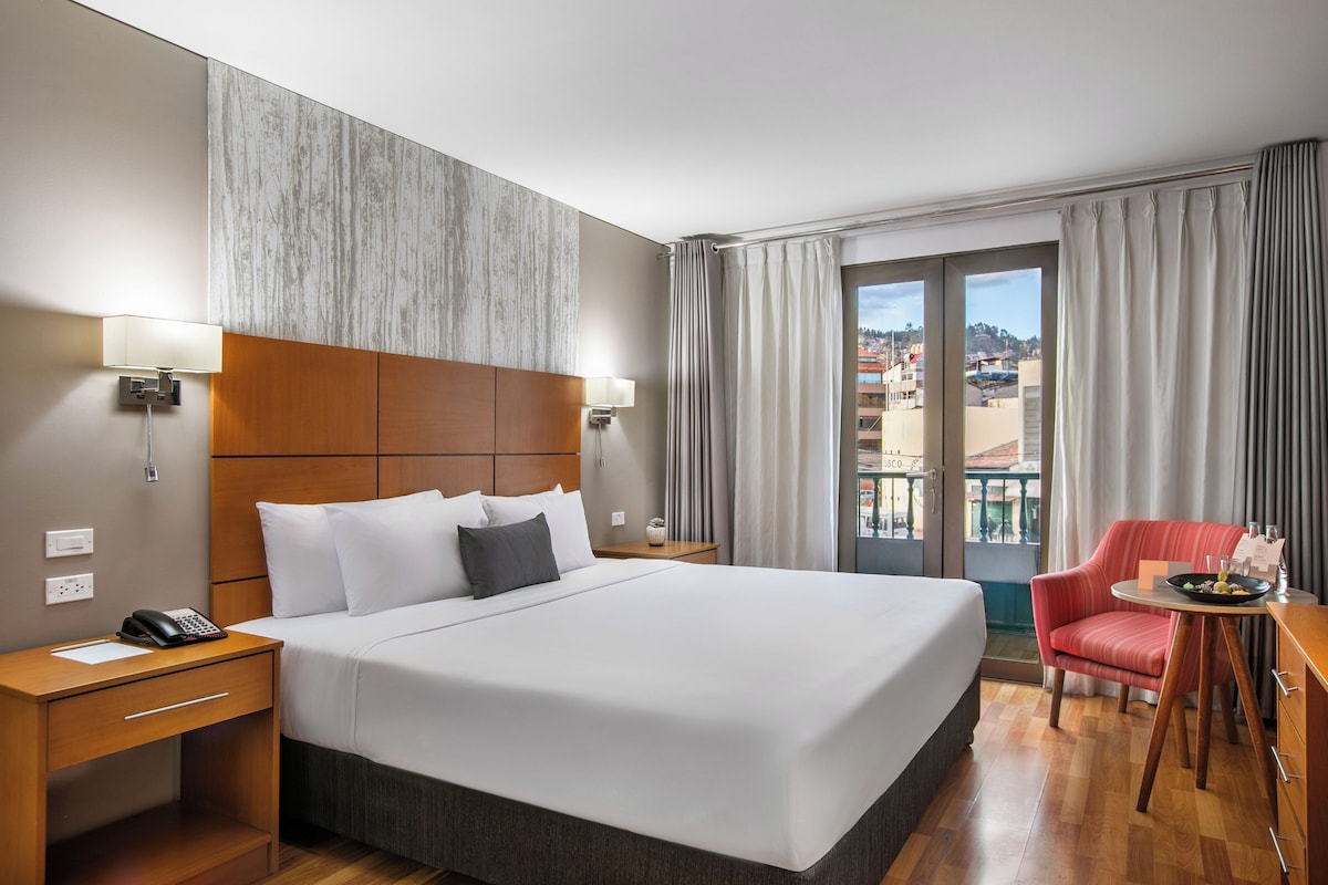 Quarto de hotel com uma cama grande, uma escrivaninha com telefone, uma cadeira e vista para a paisagem urbana pela janela.