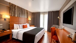 Quarto de hotel com cama grande, escrivaninha, cadeira, televisão e janela com cortinas.