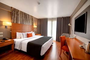 Quarto de hotel com cama grande, escrivaninha, cadeira, televisão e janela com cortinas.