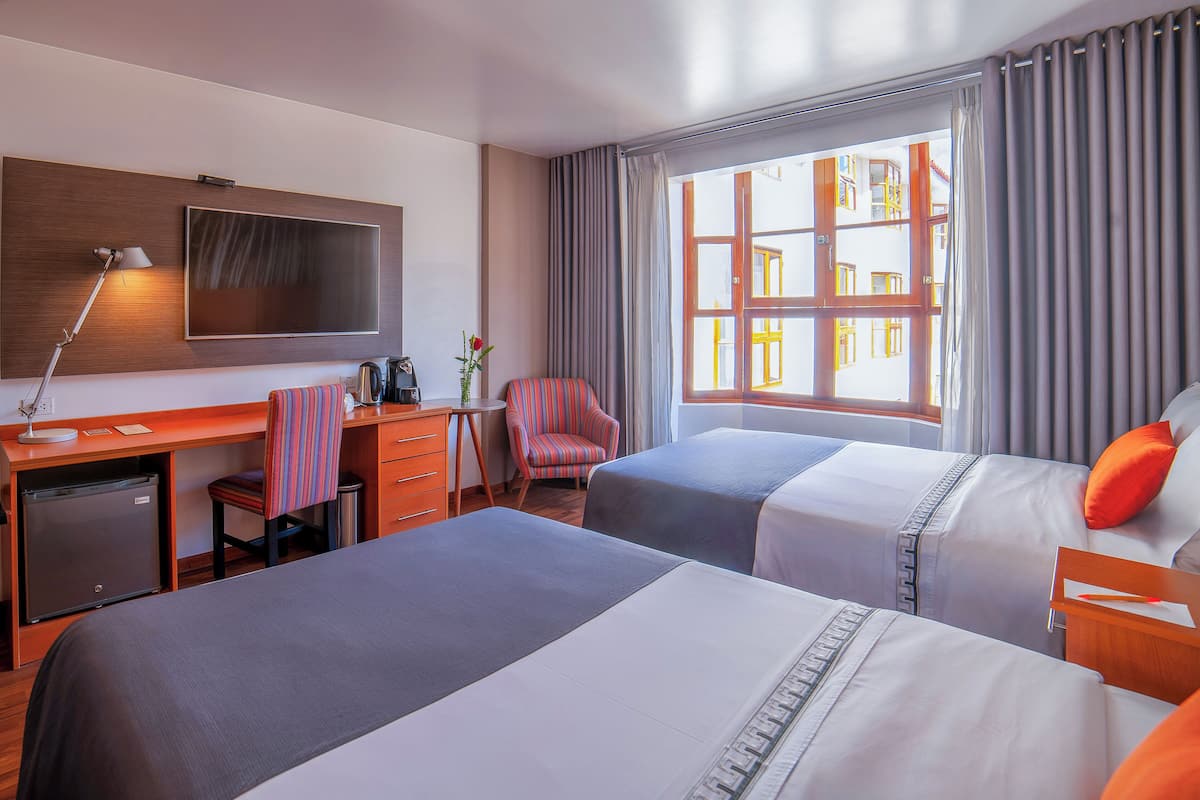Quarto de hotel com duas camas, uma secretária com computador, uma cadeira e janela com cortinas.