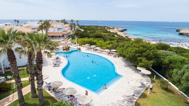 Uma piscina grande cercada por palmeiras e espreguiçadeiras, com vista para o mar e a praia ao fundo.