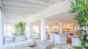 Um hall amplo com interior branco moderno, uma área de estar com sofá e pufes, e um bar com garrafas e copos.