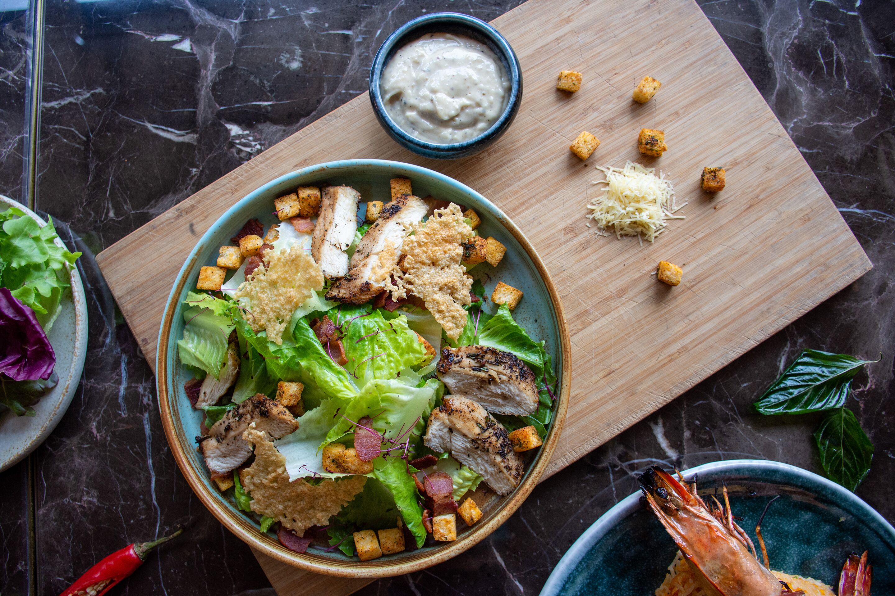 Uma saladeira com salada, croutons e queijo ralado sobre uma tábua de madeira.