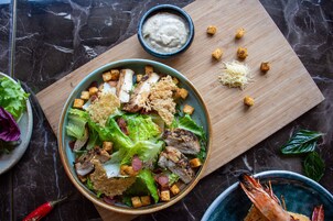 Uma saladeira com salada, croutons e queijo ralado sobre uma tábua de madeira.