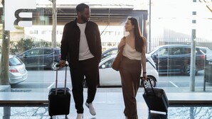 Um homem e uma mulher caminham por uma porta de vidro, carregando malas, com um estacionamento e carros visíveis ao fundo.