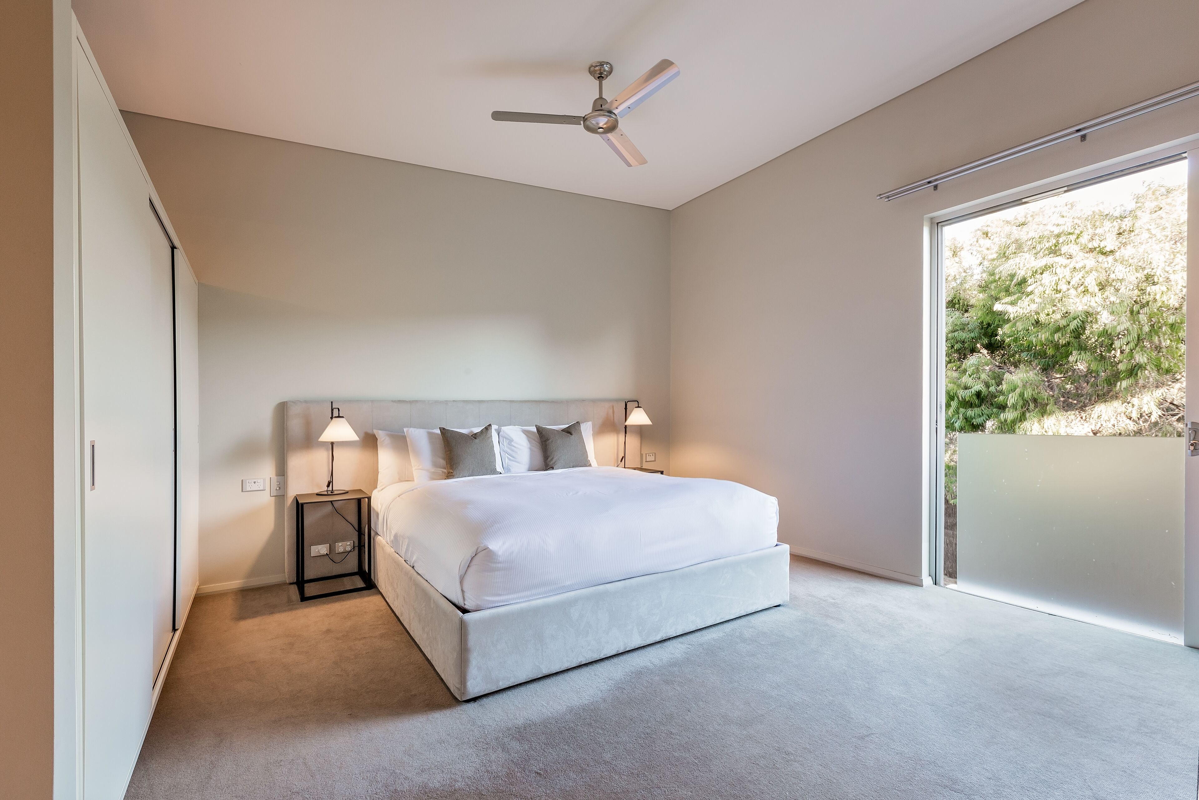 Quarto com uma cama grande, um ventilador de teto, abajures e vista para o verde através de uma porta de correr.