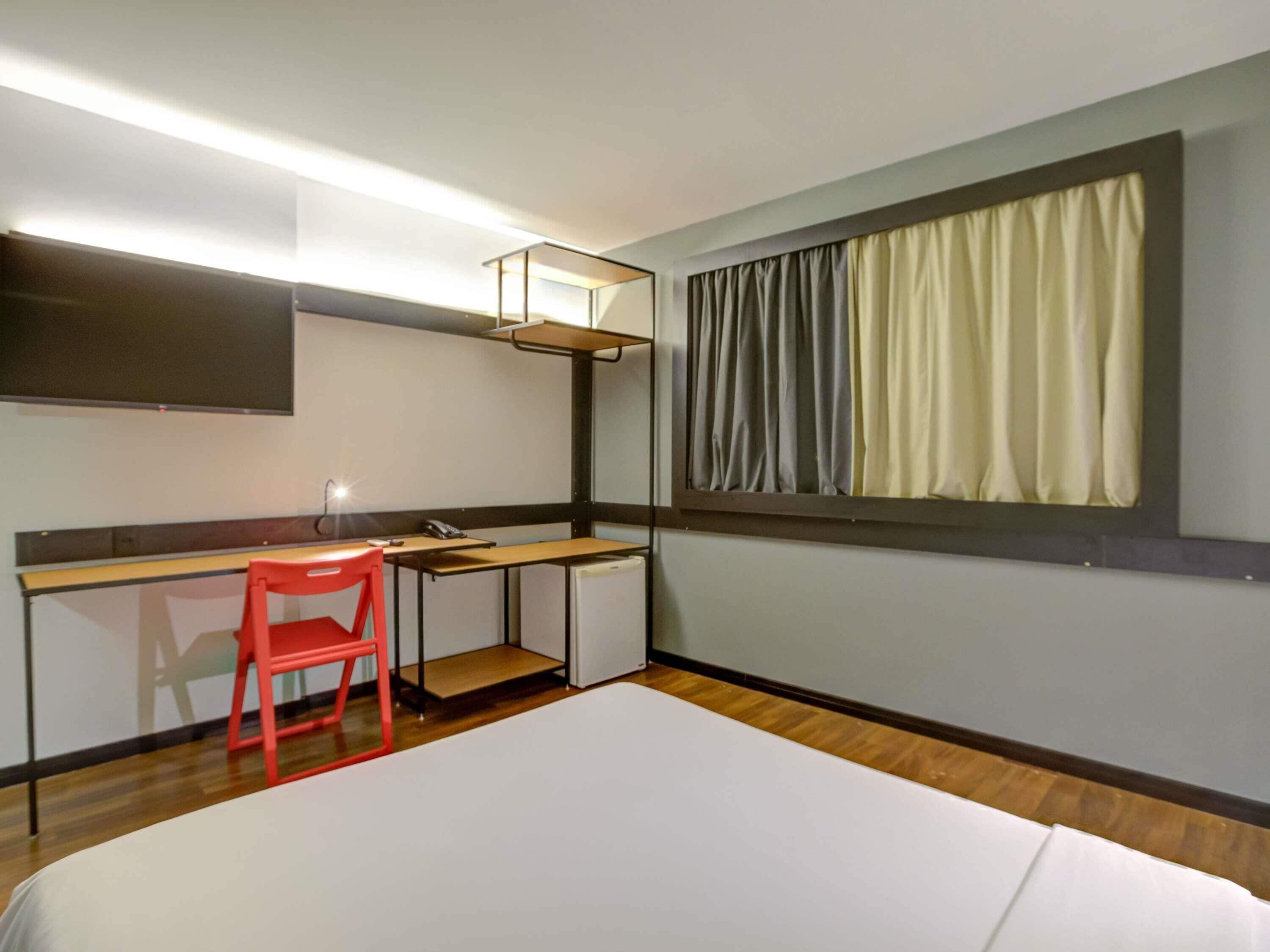 Quarto de hotel com cama, escrivaninha com cadeira, mini-geladeira e janela com cortinas.