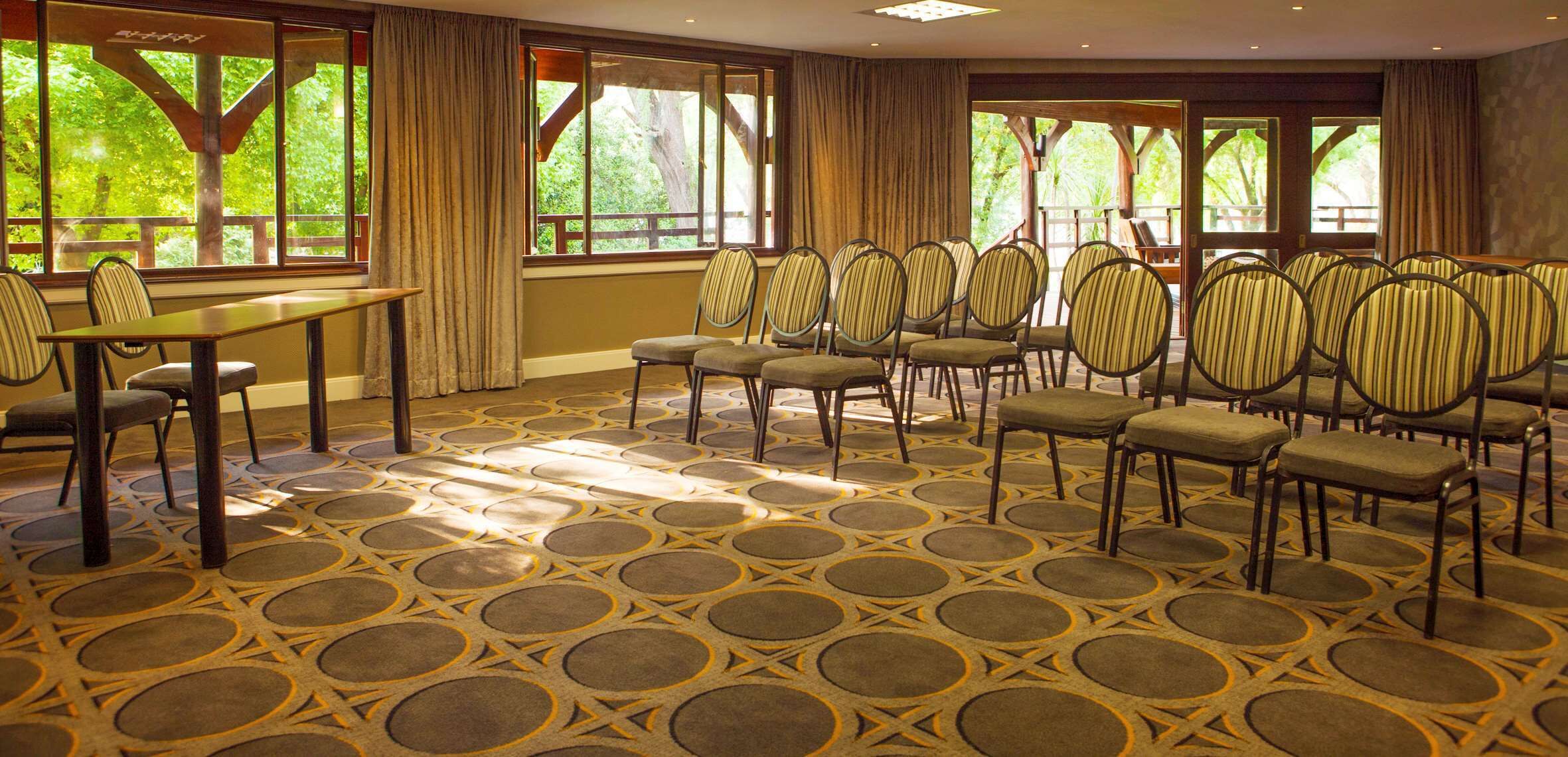 Uma sala de conferências com carpete estampado, várias cadeiras, uma mesa e janelas amplas com cortinas.