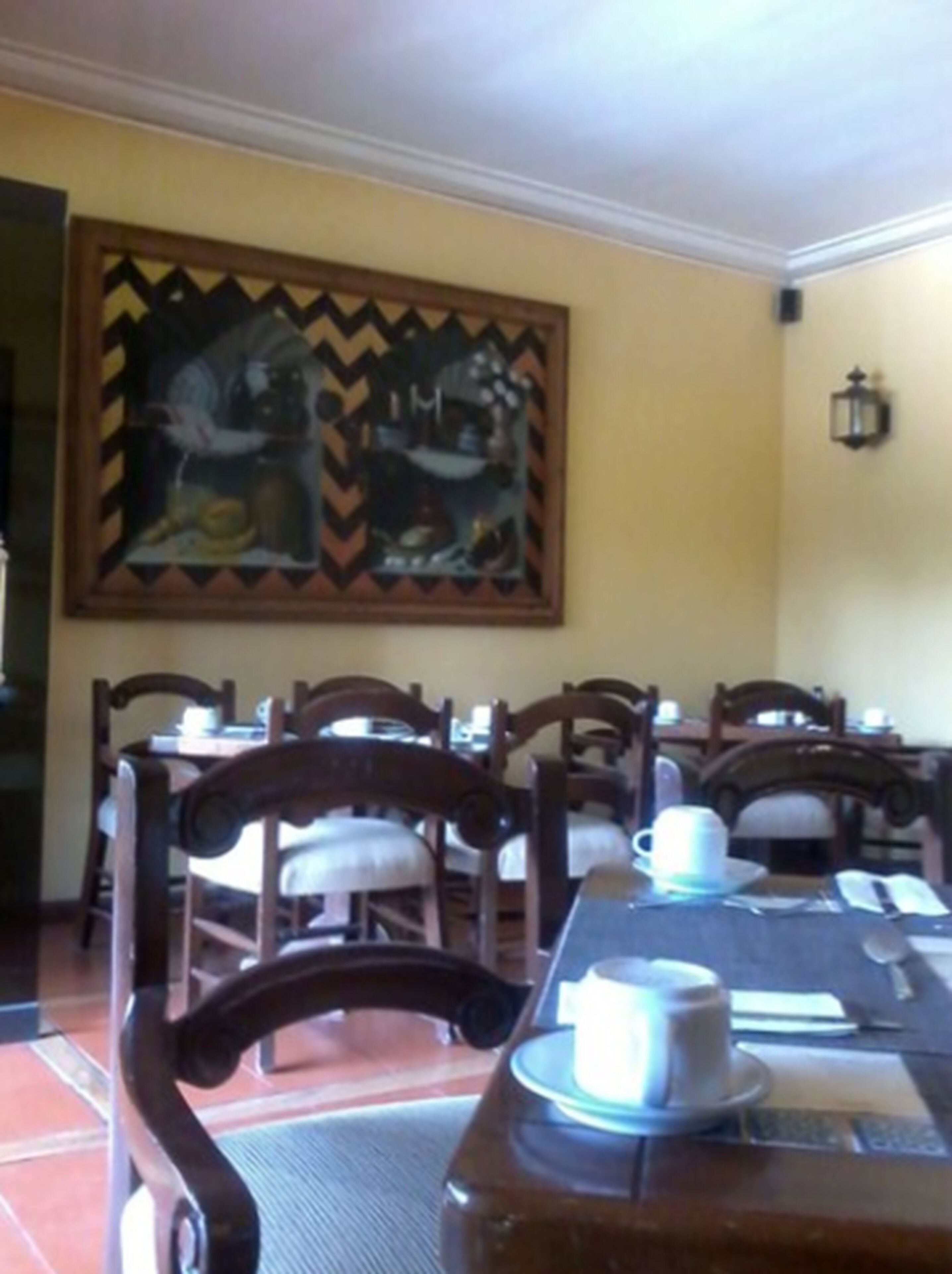 Uma área de refeições com cadeiras e mesas de madeira, uma pintura grande na parede e uma mesa com xícara e pires.