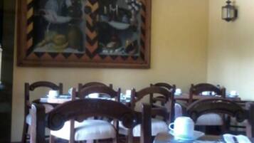 Uma área de refeições com cadeiras e mesas de madeira, uma pintura grande na parede e uma mesa com xícara e pires.