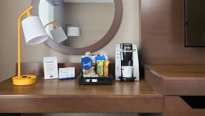 Balcão de hotel com um espelho redondo, uma luminária de mesa amarela, uma cafeteira e um tabuleiro com diversos petiscos e um cartão de boas-vindas.