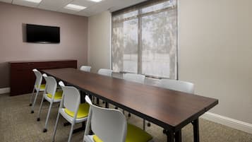 Uma sala de conferência com uma mesa comprida, cadeiras brancas com almofadas amarelas, uma TV de tela plana na parede e uma janela grande com vista para árvores.