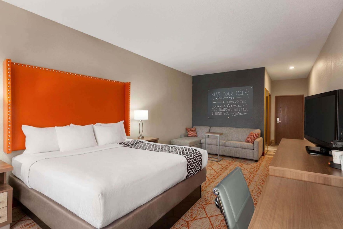 Quarto de hotel com uma cama grande, uma mesa, uma TV, um sofá e uma frase motivacional na parede.