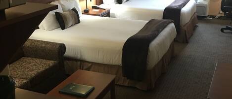 Quarto de hotel com duas camas, uma escrivaninha, uma cadeira, uma televisão e um abajur.