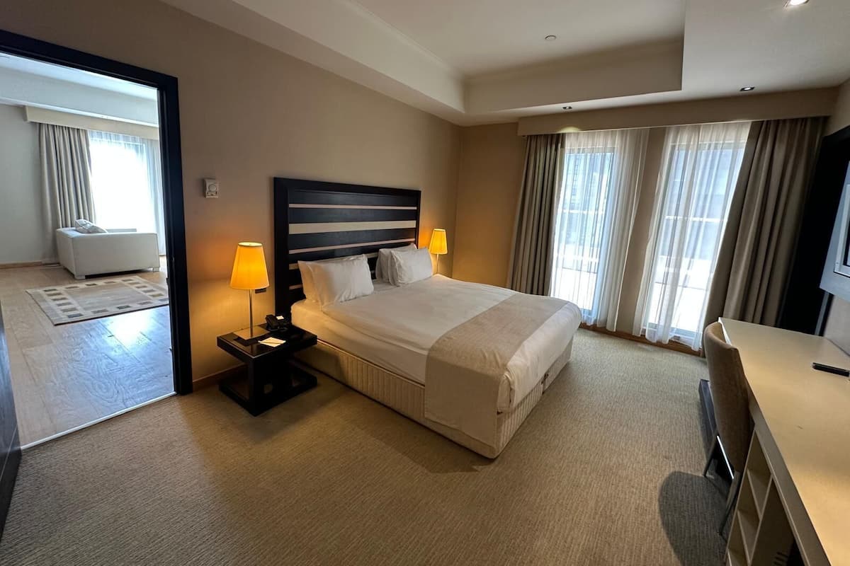Quarto de hotel com uma cama grande, luminárias de cabeceira, uma escrivaninha e vista para o exterior pela janela.
