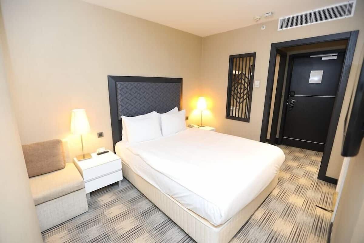 Quarto de hotel com cama, mesas de cabeceira, luminária, porta e um sistema de ventilação fixado na parede.