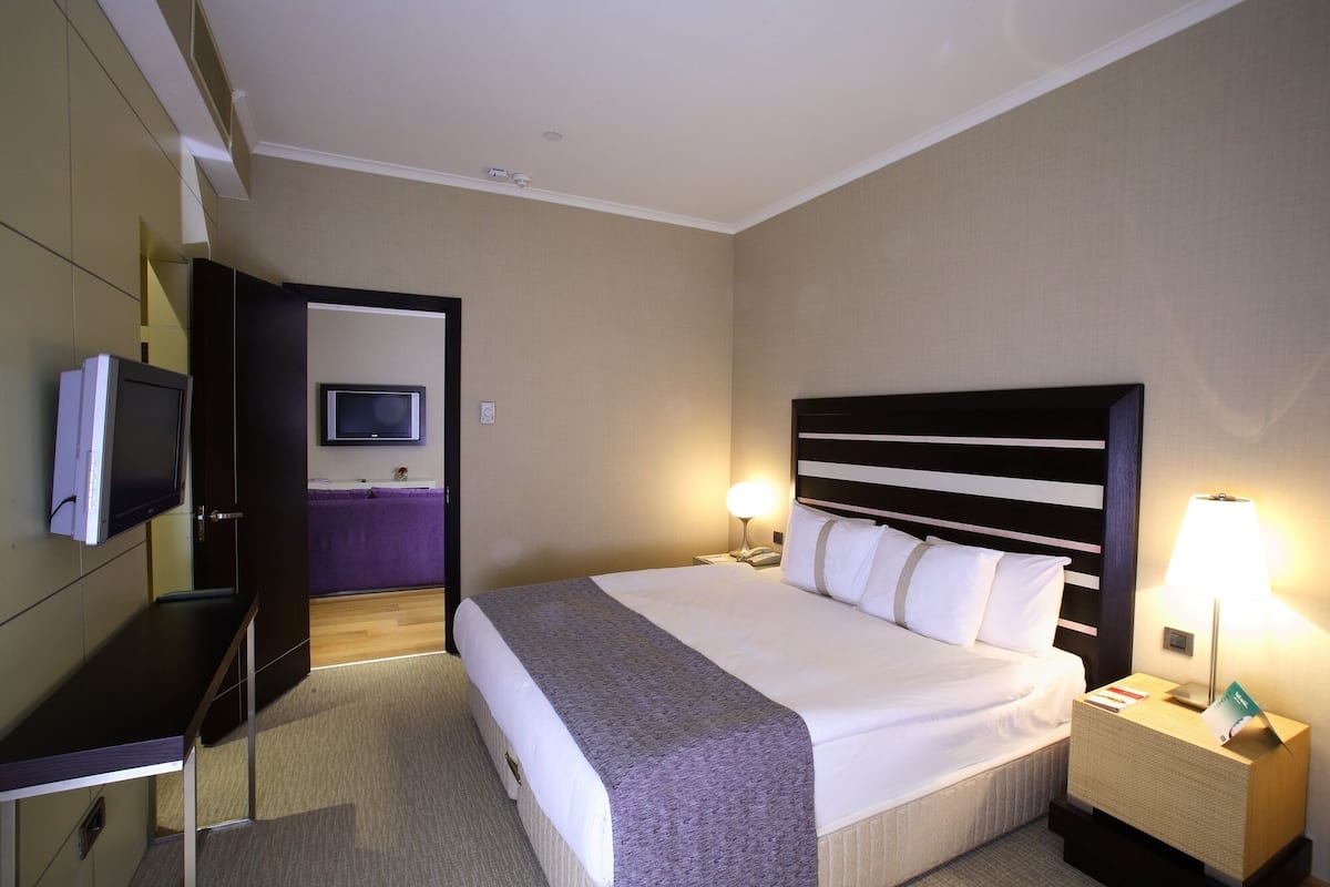 Quarto de hotel com uma cama grande, luminárias de cabeceira, uma televisão na parede e uma escrivaninha com uma cadeira.