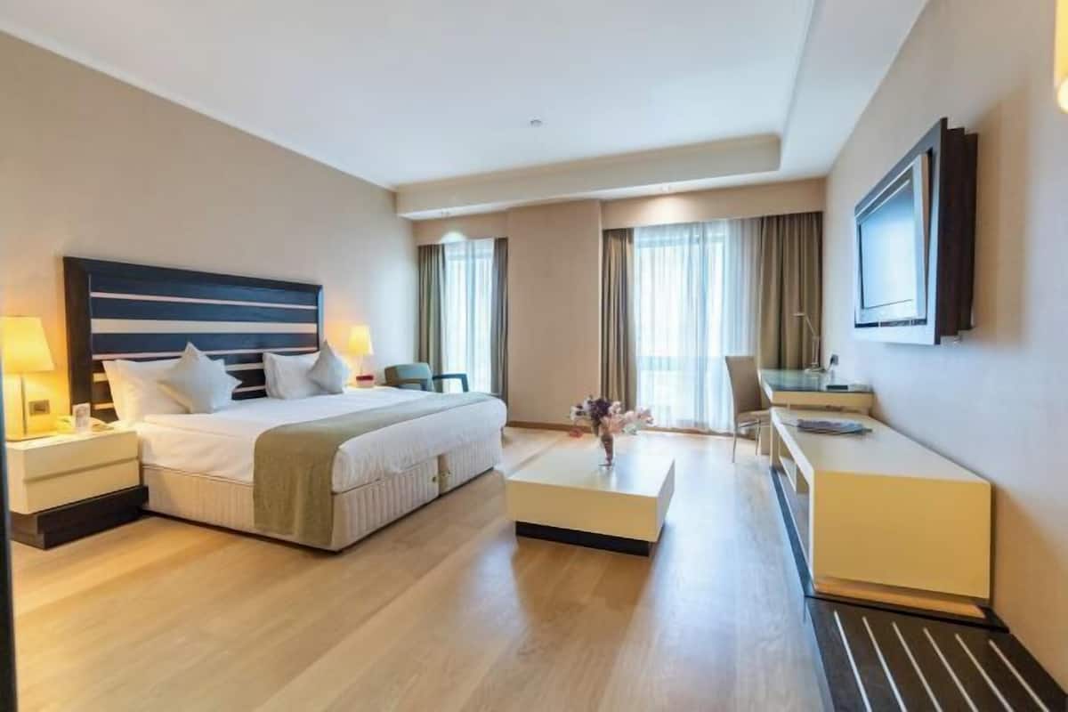 Quarto de hotel com cama, mesa de cabeceira, escrivaninha, cadeira e televisão na parede.