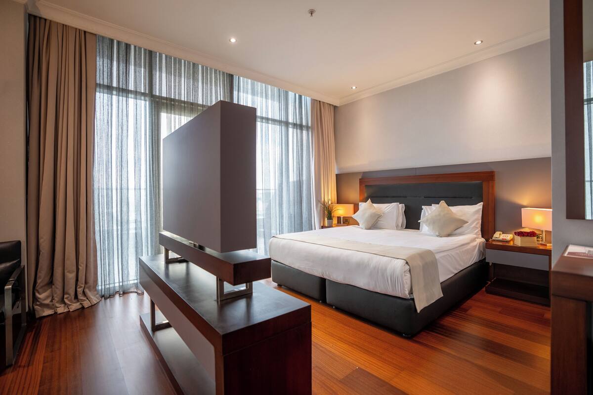 Quarto de hotel com uma cama grande, televisão, escrivaninha e mesa de cabeceira com abajur.