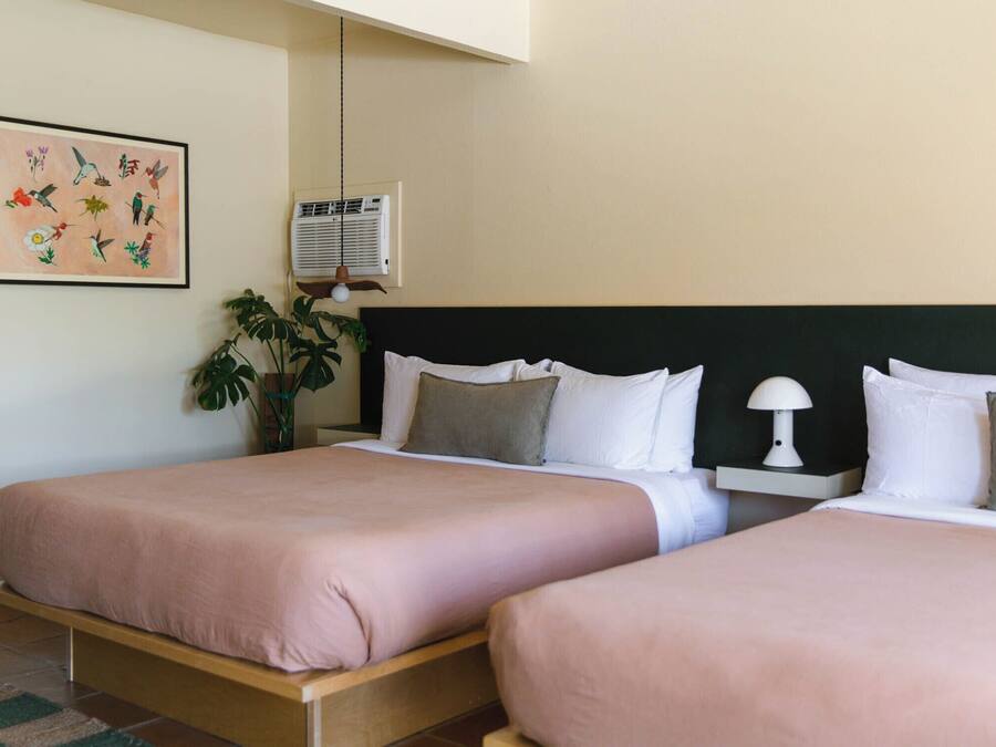 Quarto de hotel com duas camas, uma mesa de cabeceira com abajur, ar condicionado fixado na parede e uma obra de arte floral emoldurada.