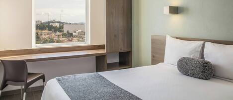 Quarto de hotel com uma janela grande, uma cama com roupa de cama branca, uma cabeceira de madeira, uma escrivaninha e uma cadeira.