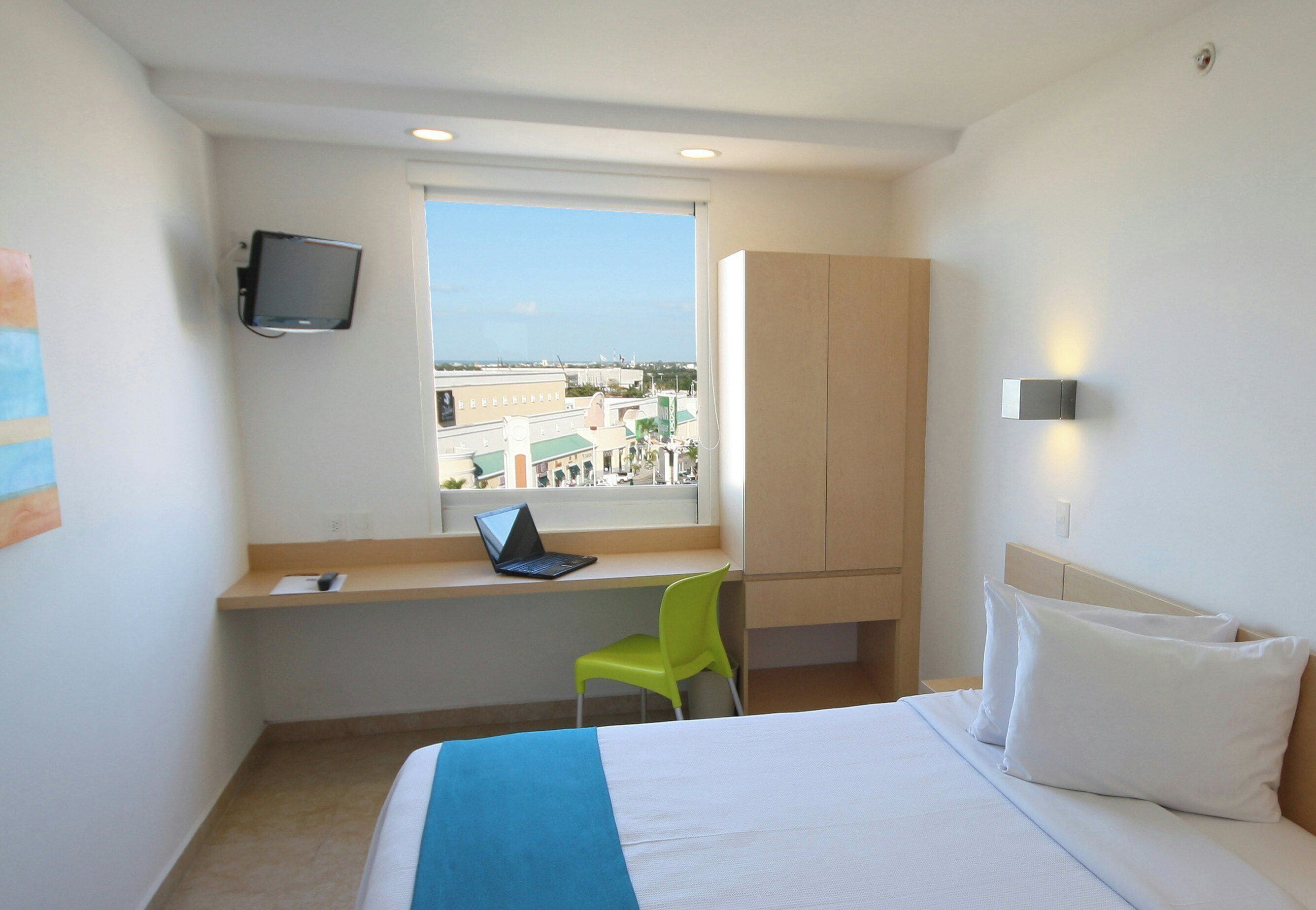 Quarto de hotel com cama, secretária com um laptop, cadeira verde, televisão na parede e janela com vista para edifícios.